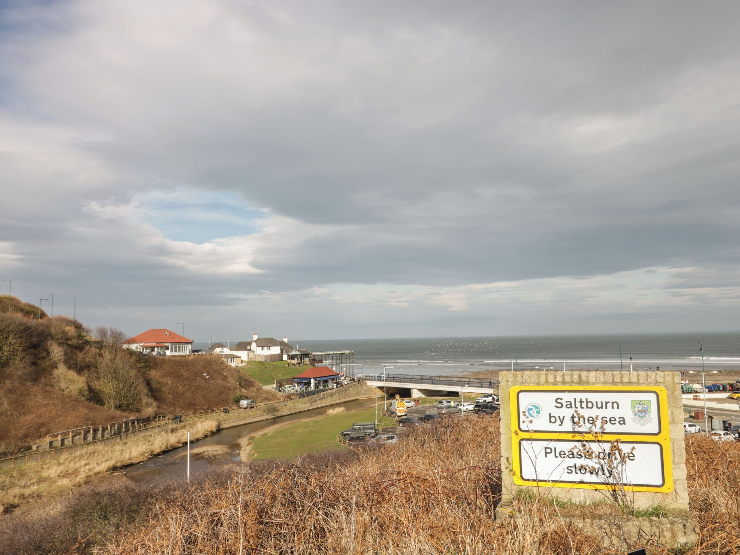 Placa de estrada costeira indicando Saltburn by the sea, com pedido para dirigir devagar.