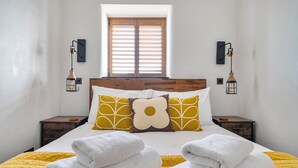 Cama bem arrumada com roupa de cama branca e amarela, duas mesas de cabeceira, uma janela com persianas de madeira e luminárias fixadas na parede.