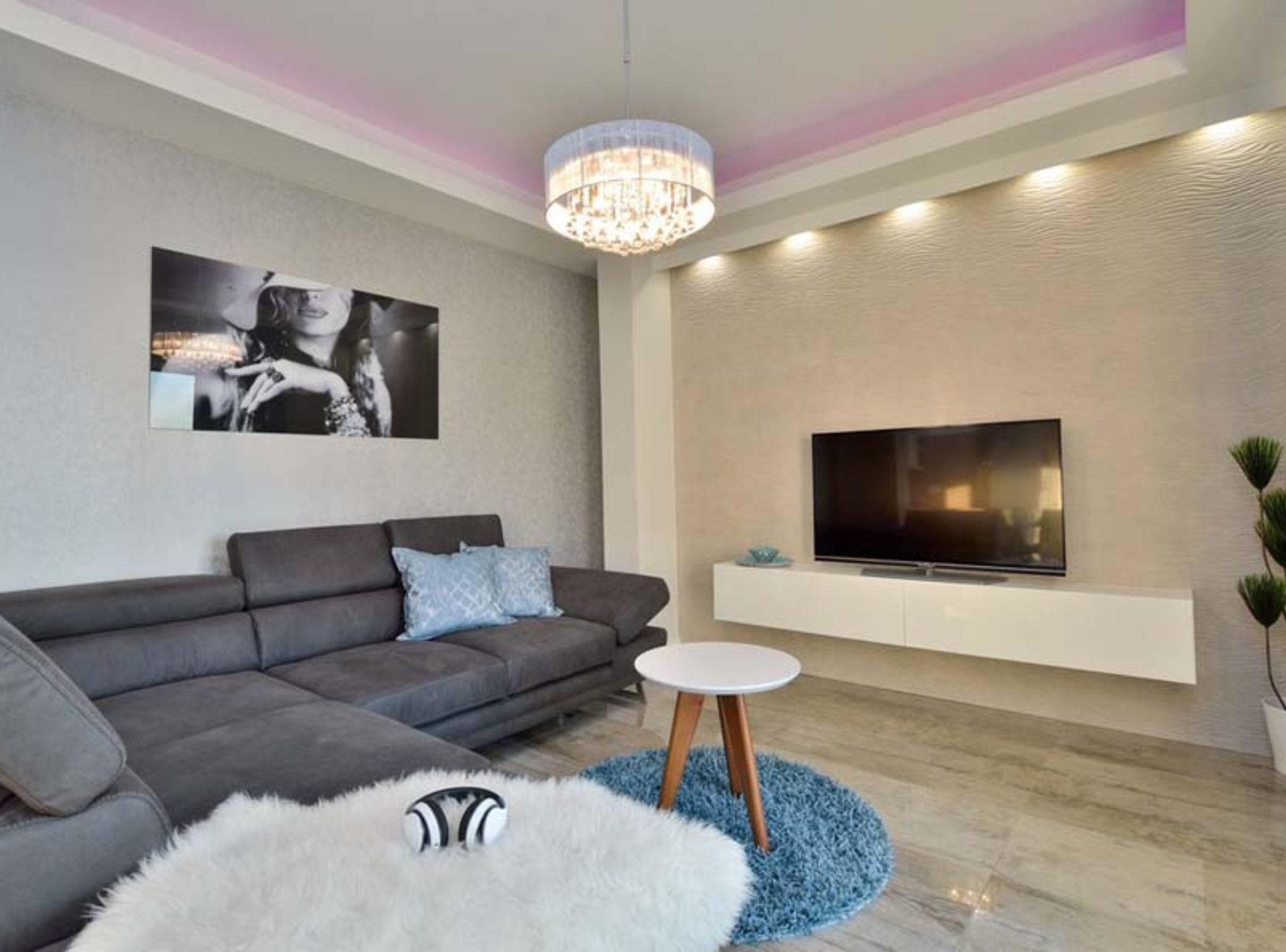 Uma sala de estar moderna com um sofá cinzento, televisão na parede, candelabro e tapete azul.