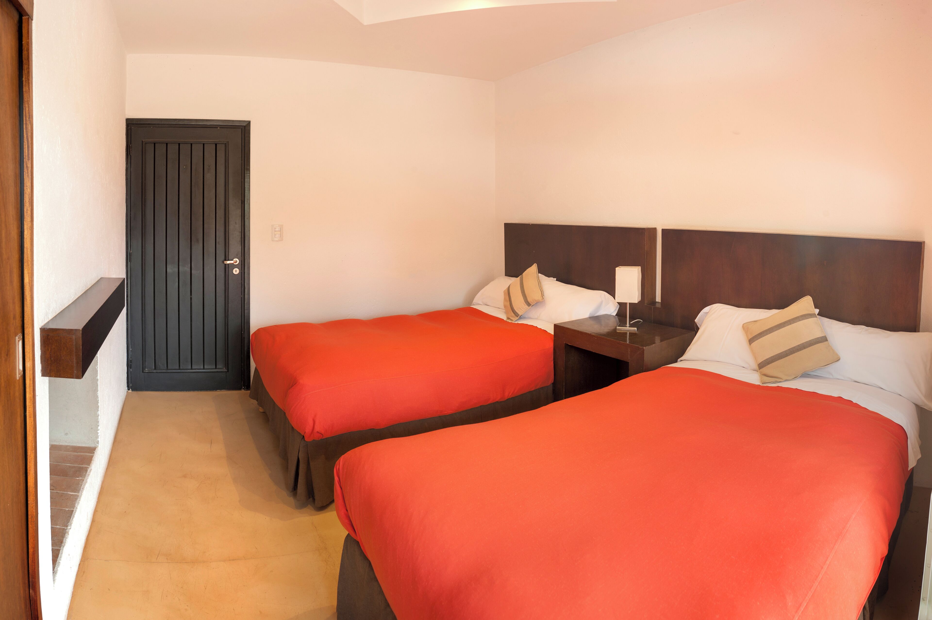 Quarto de hotel com duas camas, uma porta escura e uma mesa de cabeceira com abajur.