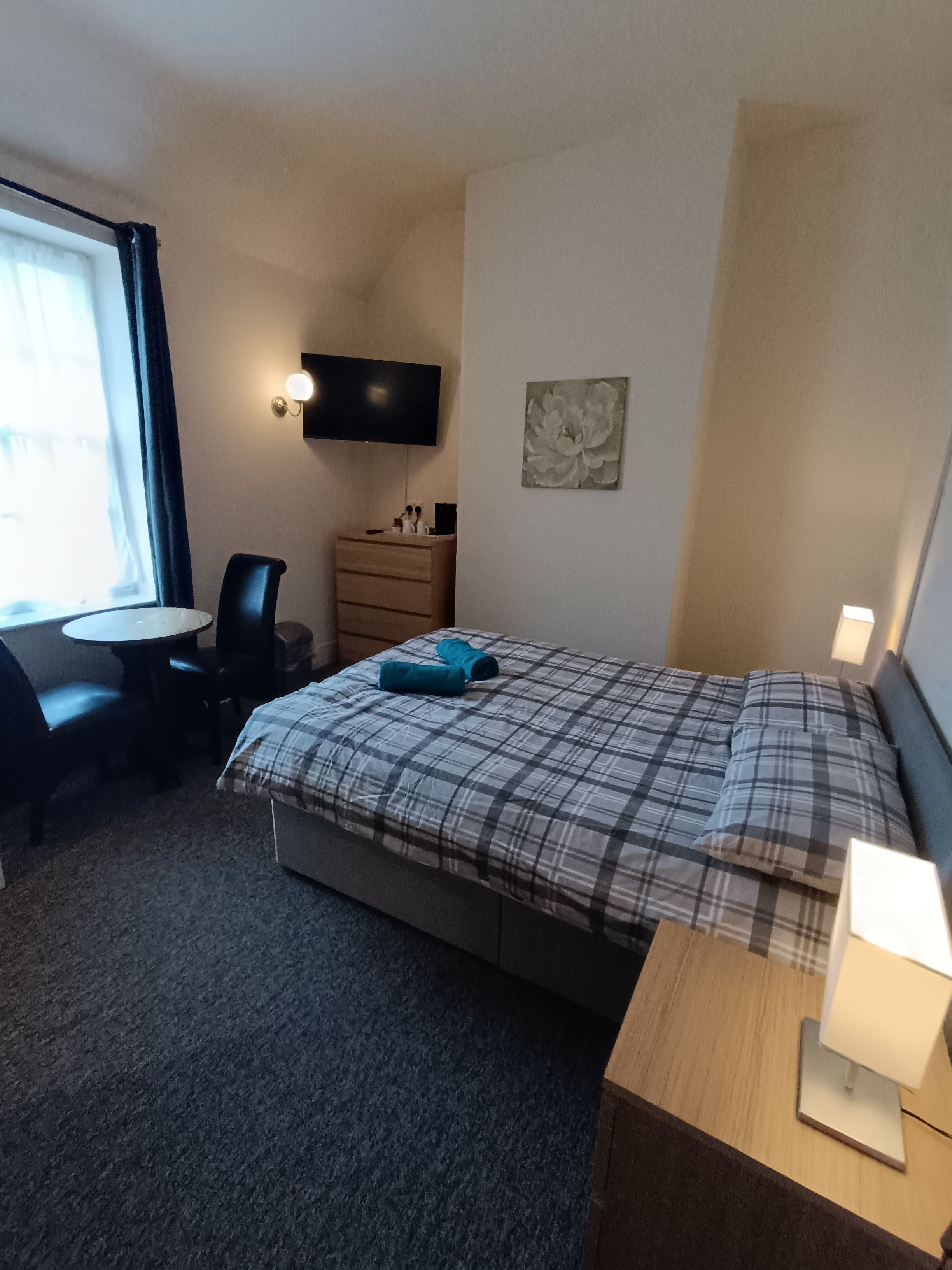 Quarto de hotel com cama, escrivaninha, poltrona, cômoda, televisão e janela.