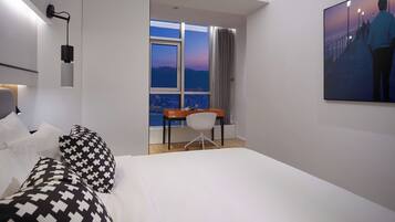 Quarto de hotel moderno com uma cama grande, uma escrivaninha e vista para a paisagem urbana através de uma grande janela.