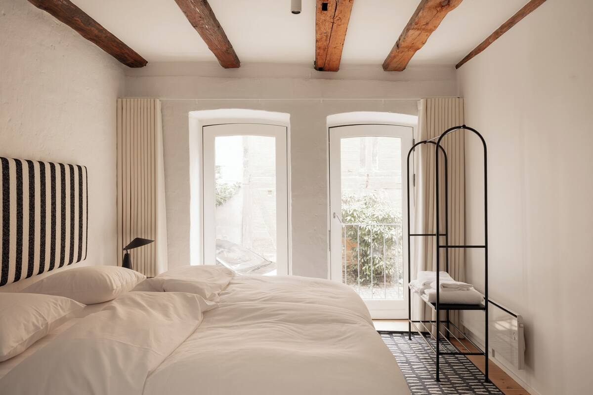 Quarto com cama, mesa de cabeceira, radiador e janela com cortinas.