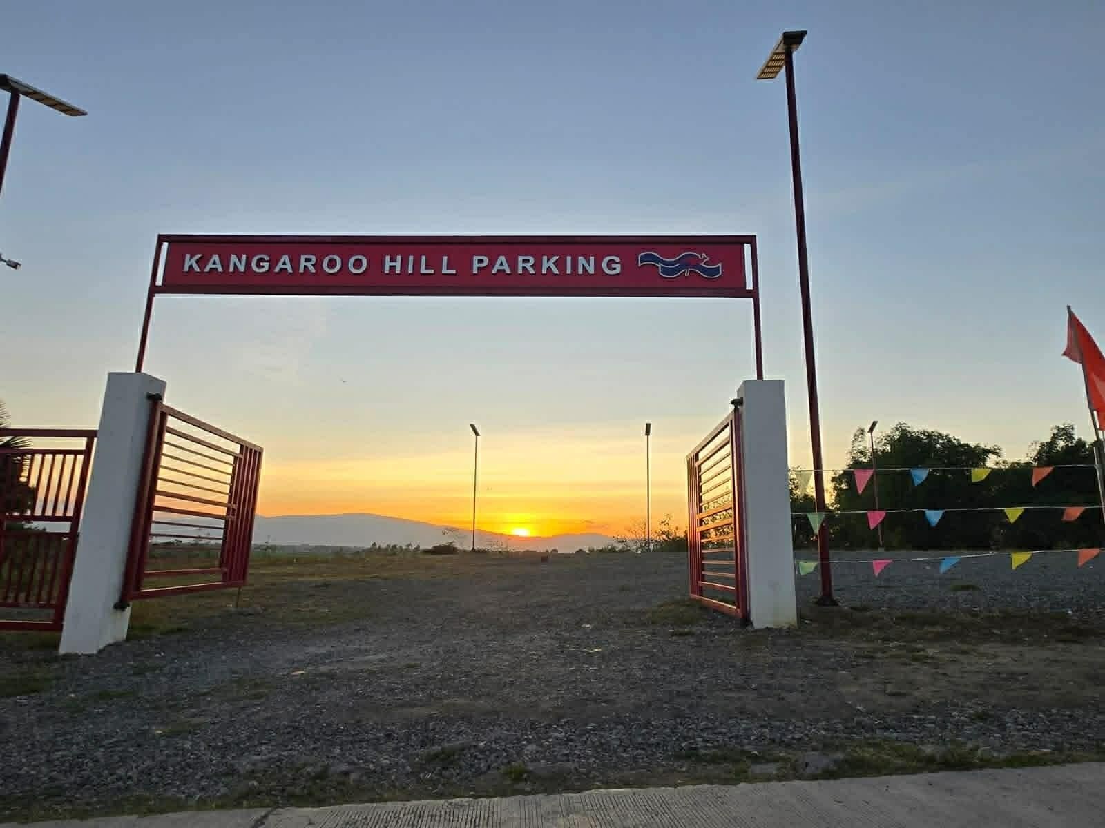 Um portão com a inscrição 'Kangaroo Hill Parking' e um pôr do sol ao fundo.