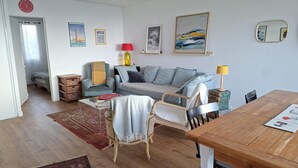 Uma sala de estar com uma mesa de jantar de madeira, um sofá, uma poltrona, um abajur e um tapete.