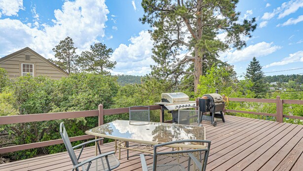 Um deck de madeira com mesa e cadeiras de vidro, uma churrasqueira e vista para uma casa e árvores.