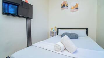 Quarto de hotel com cama, televisão de tela plana na parede e pequena mesa de cabeceira com luminária.
