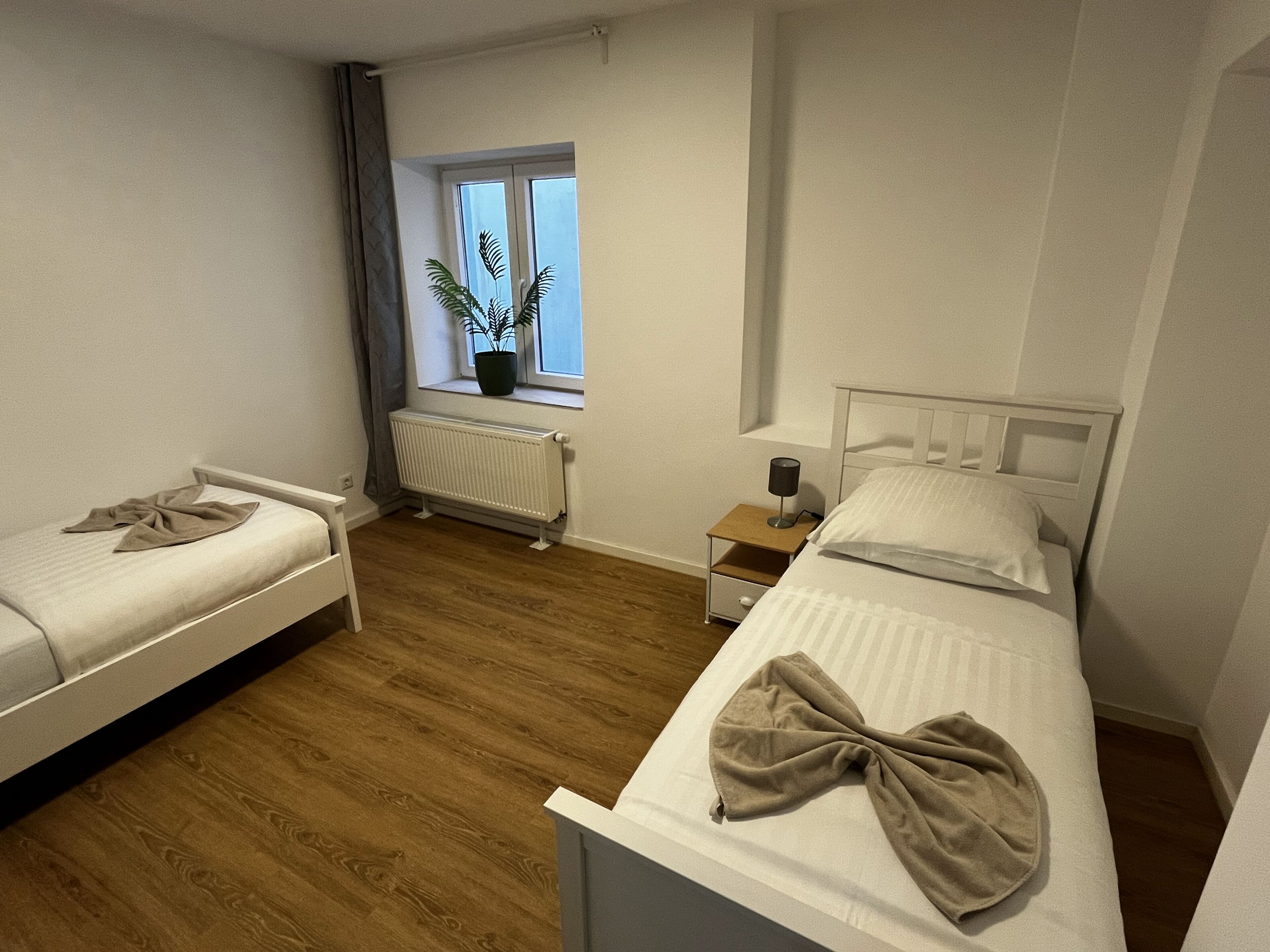 Quarto com duas camas, janela com planta, radiador e mesa de cabeceira com abajur.
