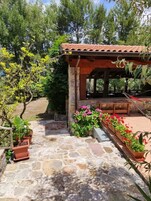 Um pátio com piso de pedra, pérgola de madeira, plantas em vasos e um caminho de pedra que leva a um edifício.