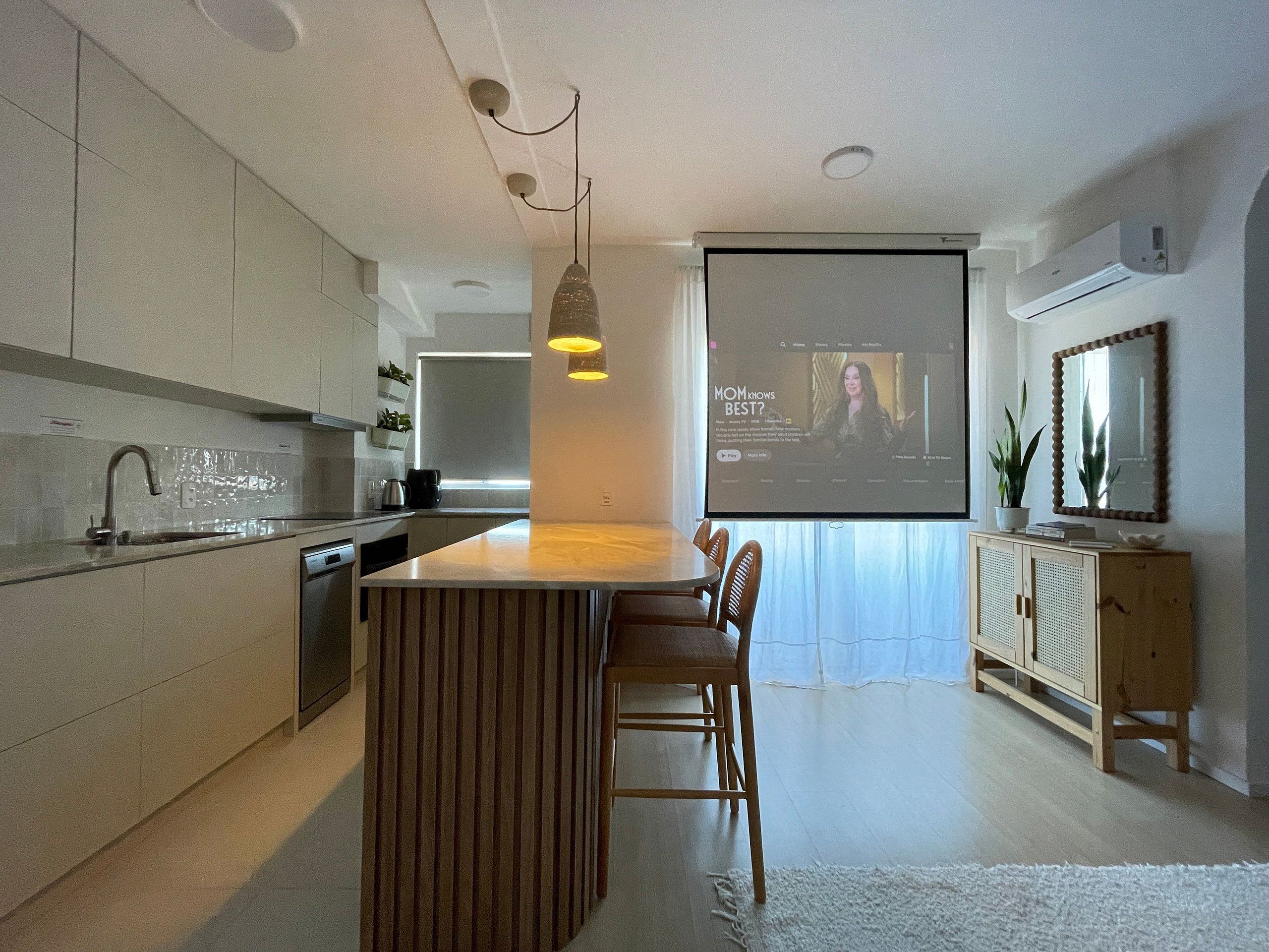 Cozinha moderna com ilha central, área de jantar com cadeiras, tela grande exibindo um vídeo e armário de madeira com objetos decorativos.
