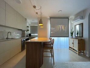 Cozinha moderna com ilha central, área de jantar com cadeiras, tela grande exibindo um vídeo e armário de madeira com objetos decorativos.