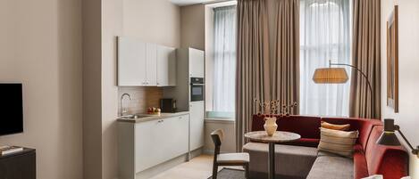 Quarto de hotel moderno com uma cama grande, um sofá, uma pequena mesa de jantar, uma área de cozinha e uma grande janela com cortinas.