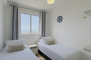 Quarto com duas camas, uma janela com cortinas, um relógio de parede e uma mesinha.