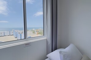 Quarto de hotel com uma janela grande, cama com roupa de cama branca e vista para o mar e edifícios.