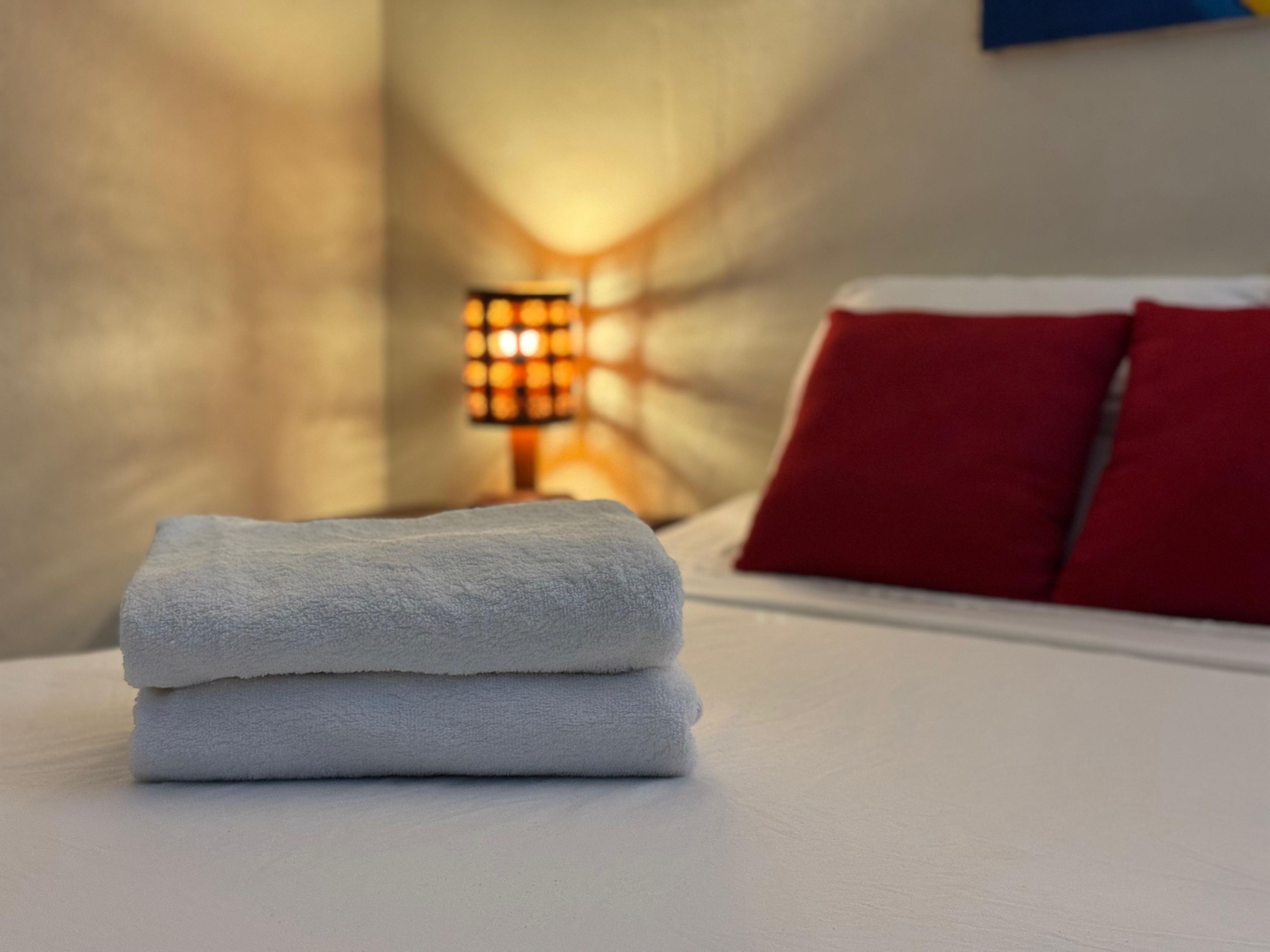 Cama bem arrumada com edredom branco e duas almofadas vermelhas, uma toalha branca dobrada e uma luminária de parede com grade de luzes.