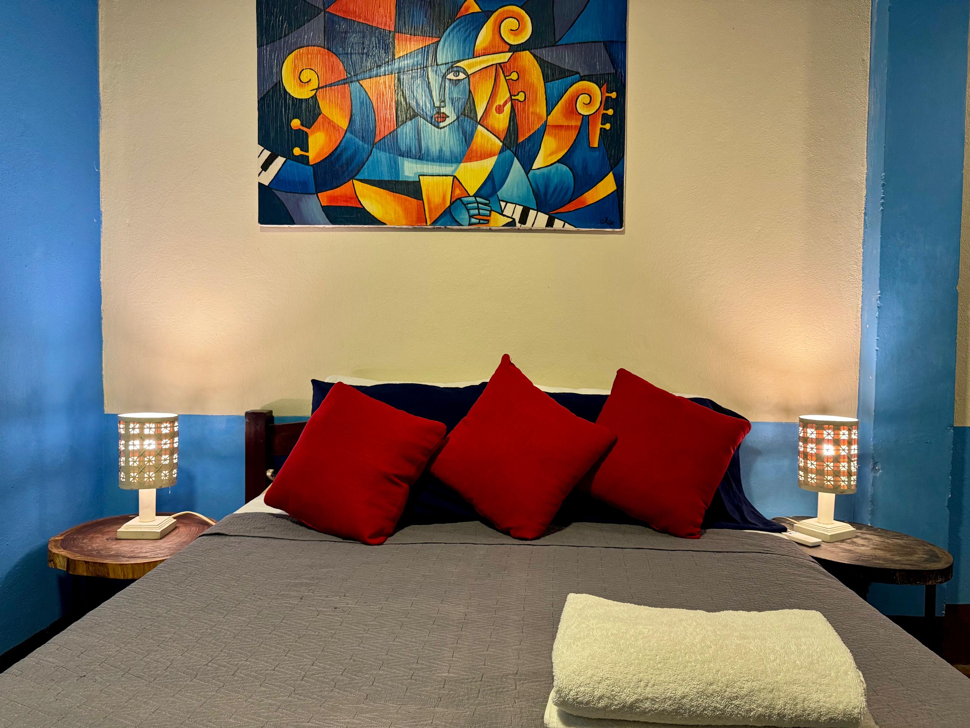 Cama com travesseiros vermelhos, manta cinza e toalha branca. A parede azul e bege tem um grande quadro abstrato acima da cama.