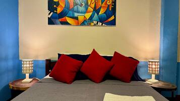 Cama com travesseiros vermelhos, manta cinza e toalha branca. A parede azul e bege tem um grande quadro abstrato acima da cama.
