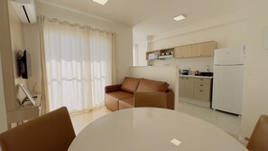 Um espaço de estar moderno e compacto, com área de refeições, sofá, cozinha com geladeira e janela com cortinas.
