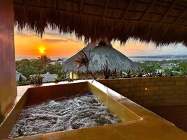 Uma banheira de hidromassagem com vista para um pôr do sol e uma construção com telhado de palha.