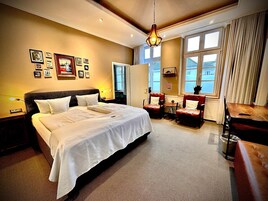 Quarto de hotel com uma cama grande, uma escrivaninha, uma cadeira, uma mesinha e uma janela com cortinas.