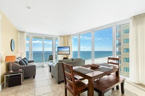 Ampla sala de estar com uma grande janela com vista para o oceano, uma mesa de jantar com cadeiras, um sofá e uma televisão na parede.