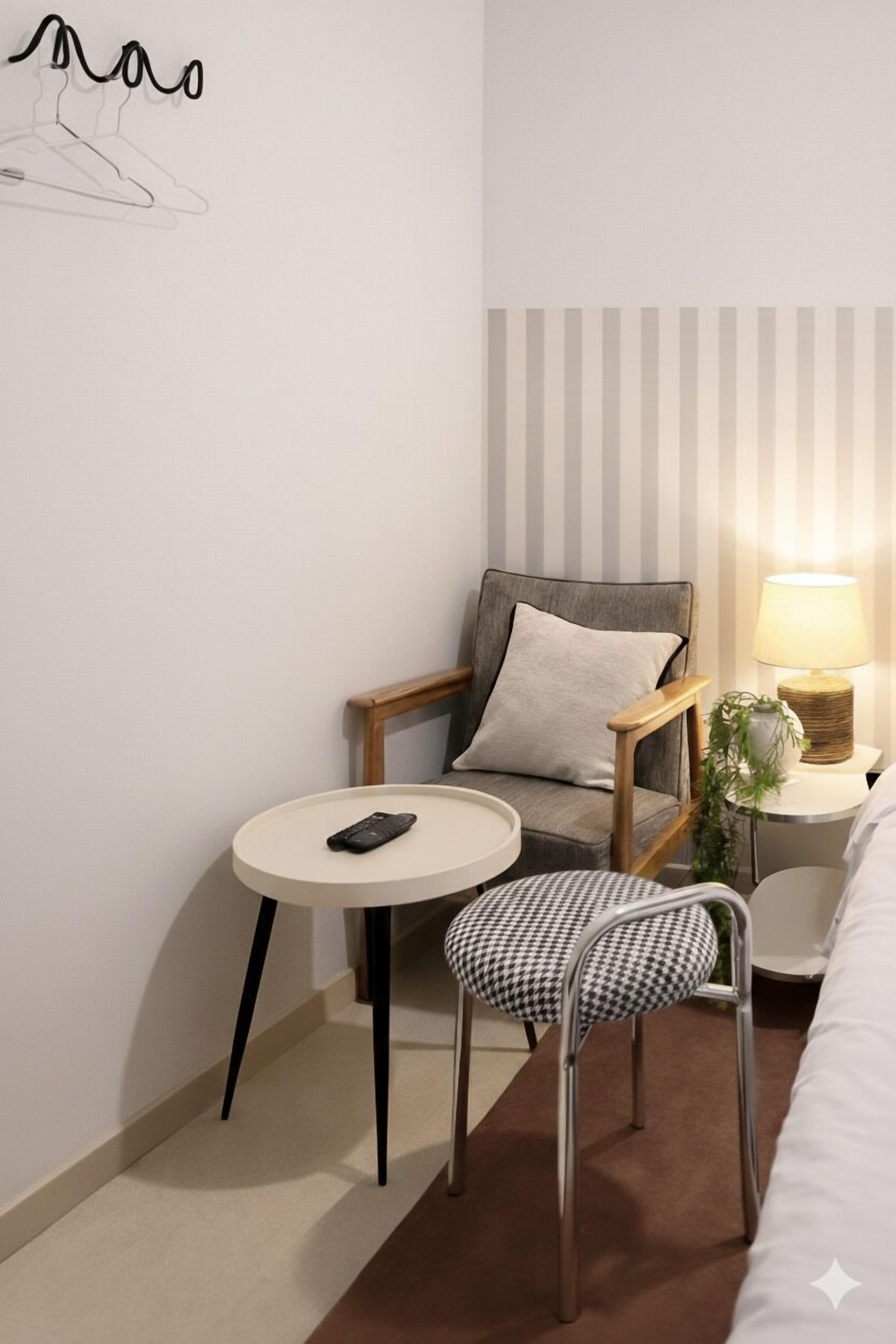 Quarto de hotel pequeno e bem organizado, com uma cama de solteiro, uma cadeira, uma mesinha e um abajur.
