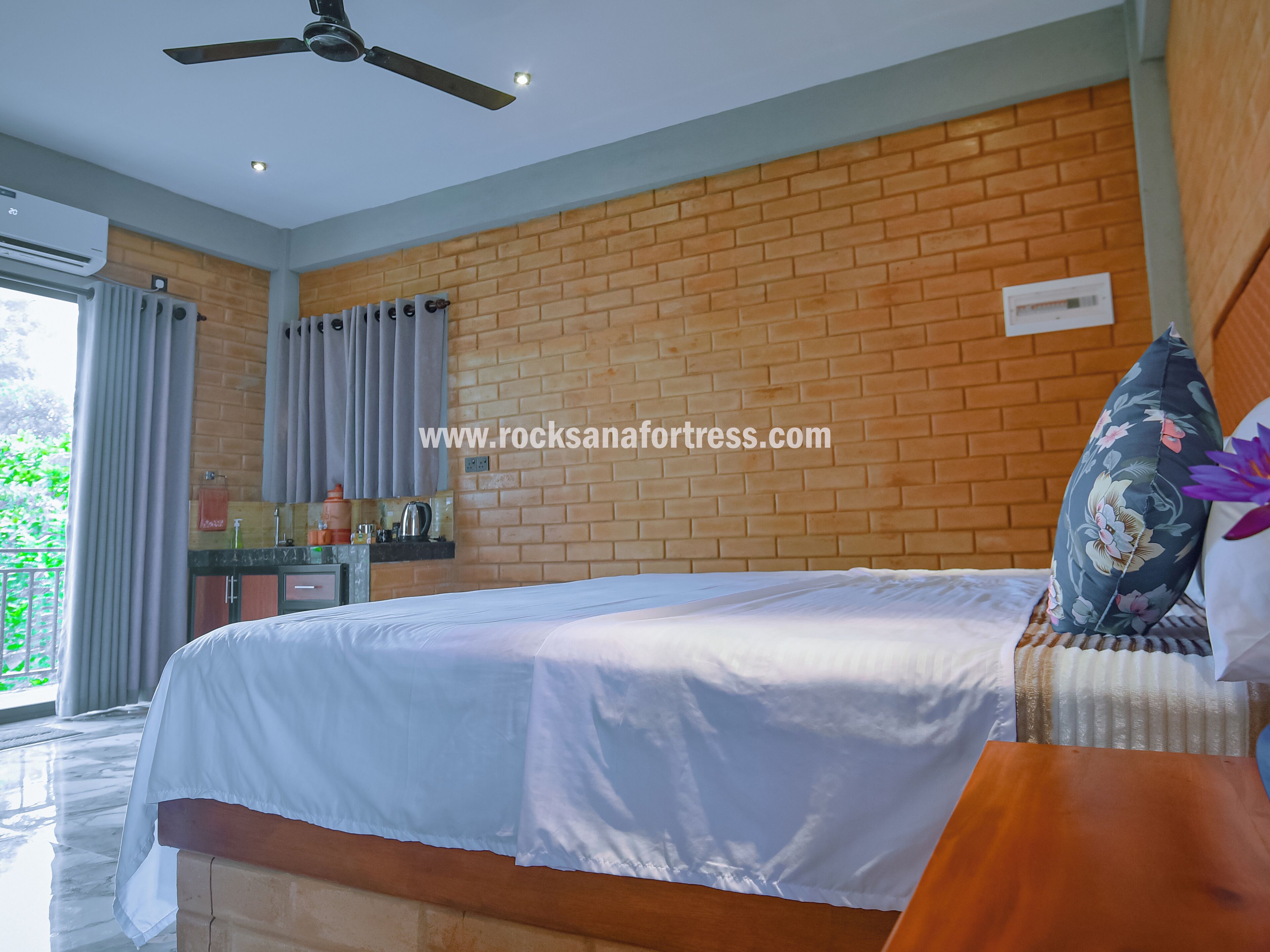 Quarto de hotel com parede de tijolos, cama com roupa de cama branca, ventilador de teto e vista para o verde lá fora.