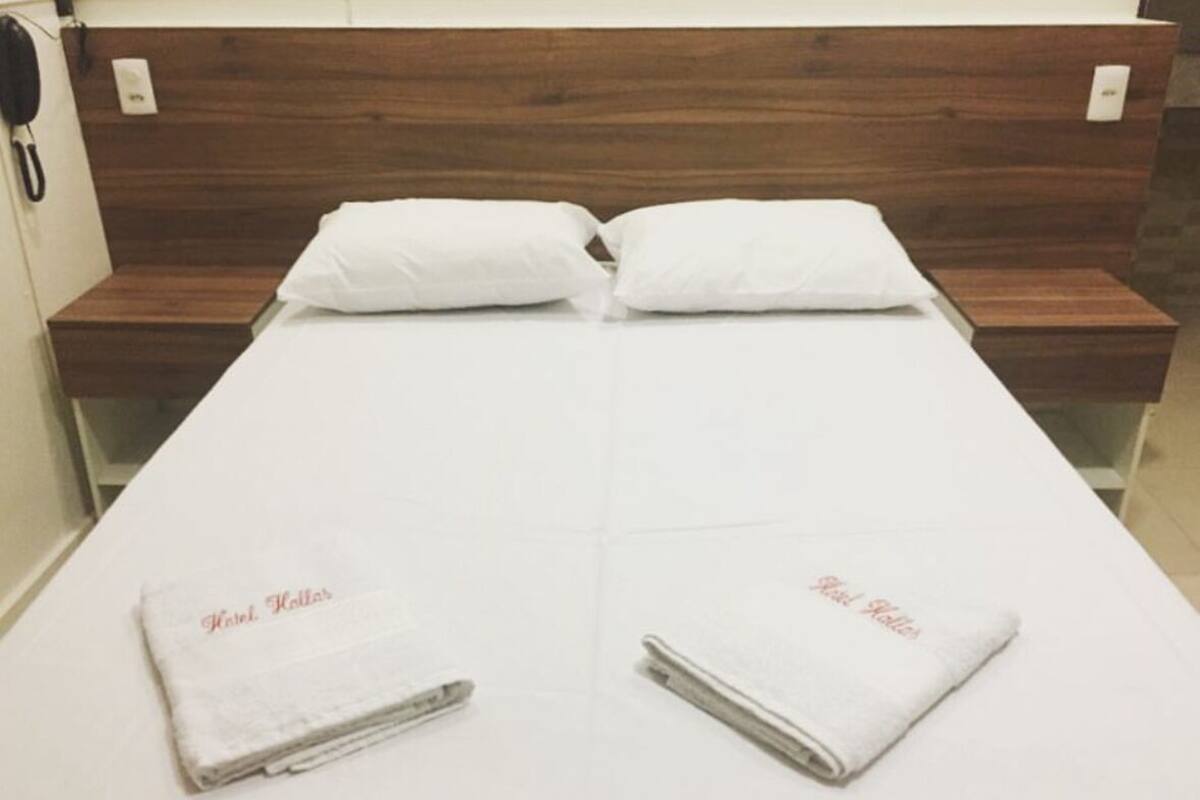 Quarto de hotel com cabeceira de madeira, dois travesseiros brancos e duas toalhas bordadas com 'Hotel Keller'.