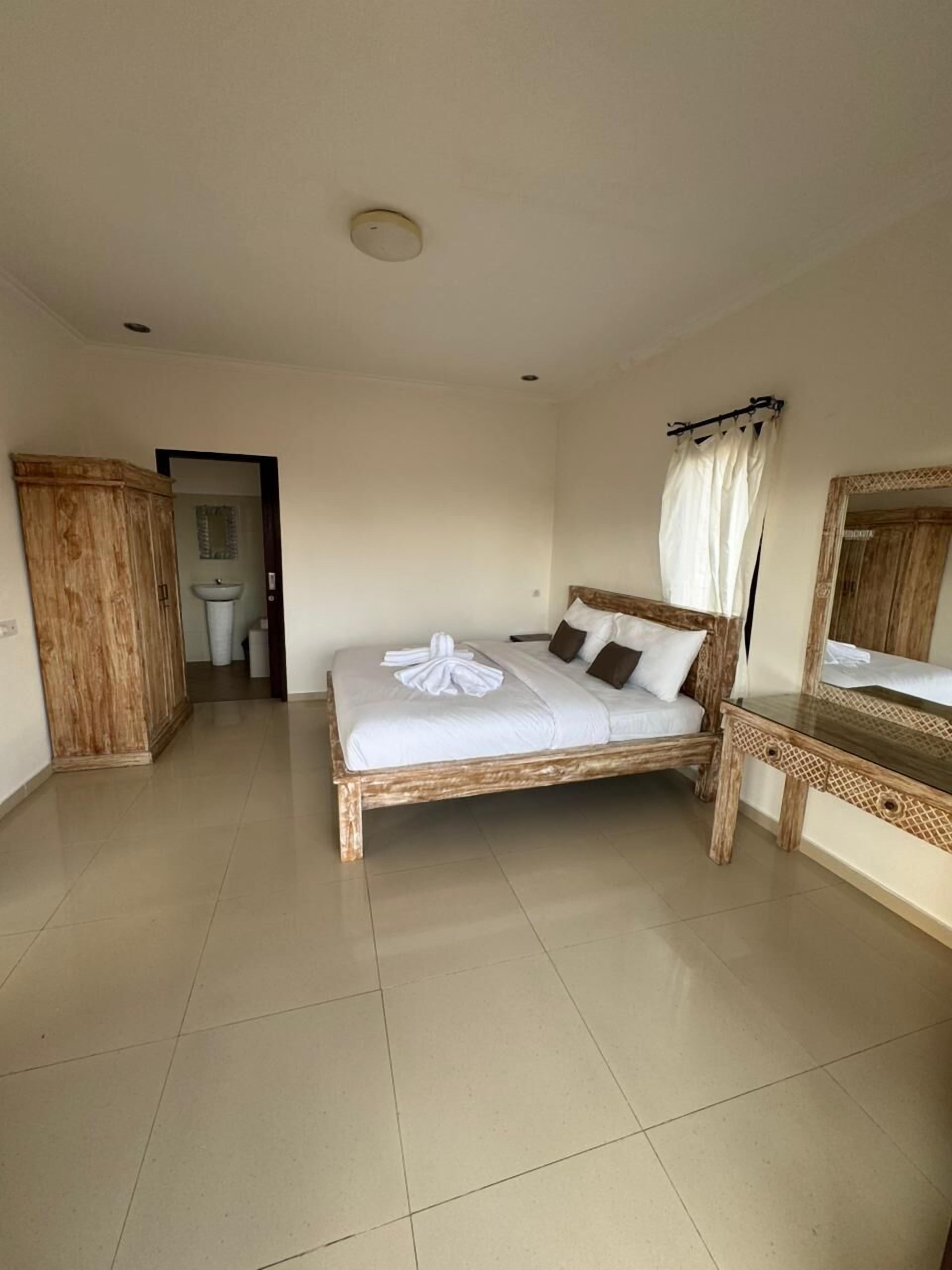 Quarto com cama, guarda-roupa de madeira, mesa de cabeceira e banheiro visível através de uma porta aberta.
