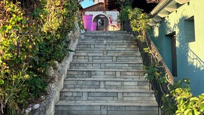 Uma escadaria de pedra leva a um edifício com varanda, cercado por vegetação e um céu claro.