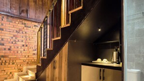 Uma escadaria com degraus de madeira e um piso com padrão geométrico, que leva a uma área de cozinha com chaleira de aço inoxidável e armários.