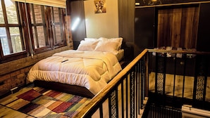 Quarto aconchegante com teto de madeira, cama com colcha, janela com persianas e escada com corrimão.