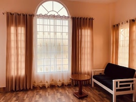 Quarto com uma grande janela em arco, cortinas bege, uma mesa redonda de madeira e um sofá preto.