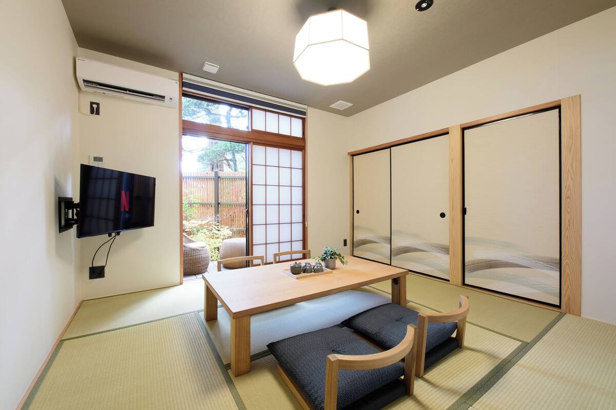 Um quarto japonês tradicional com tatames, uma mesa baixa com almofadas, televisão, portas deslizantes e vista para um jardim.