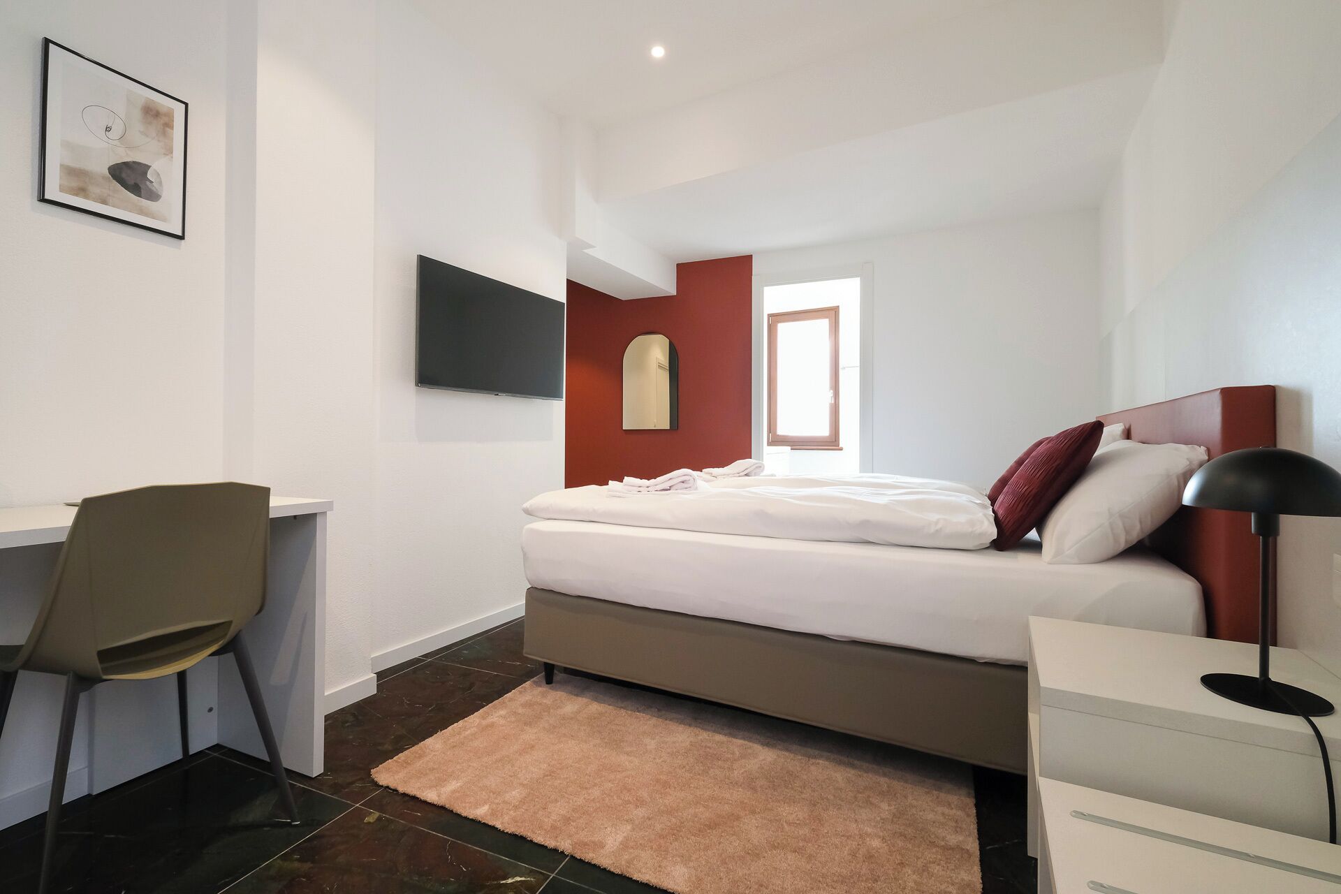 Quarto de hotel moderno com cama, escrivaninha, cadeira e televisão.