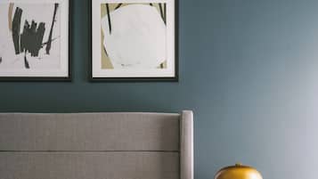 Uma cama bem arrumada com lençóis brancos, uma cabeceira estofada cinza, uma mesa de cabeceira com abajur dourado e duas obras de arte abstratas emolduradas na parede.