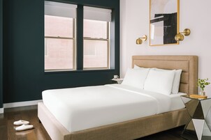 Quarto com cama, mesa de cabeceira, janela e espelho fixado na parede.