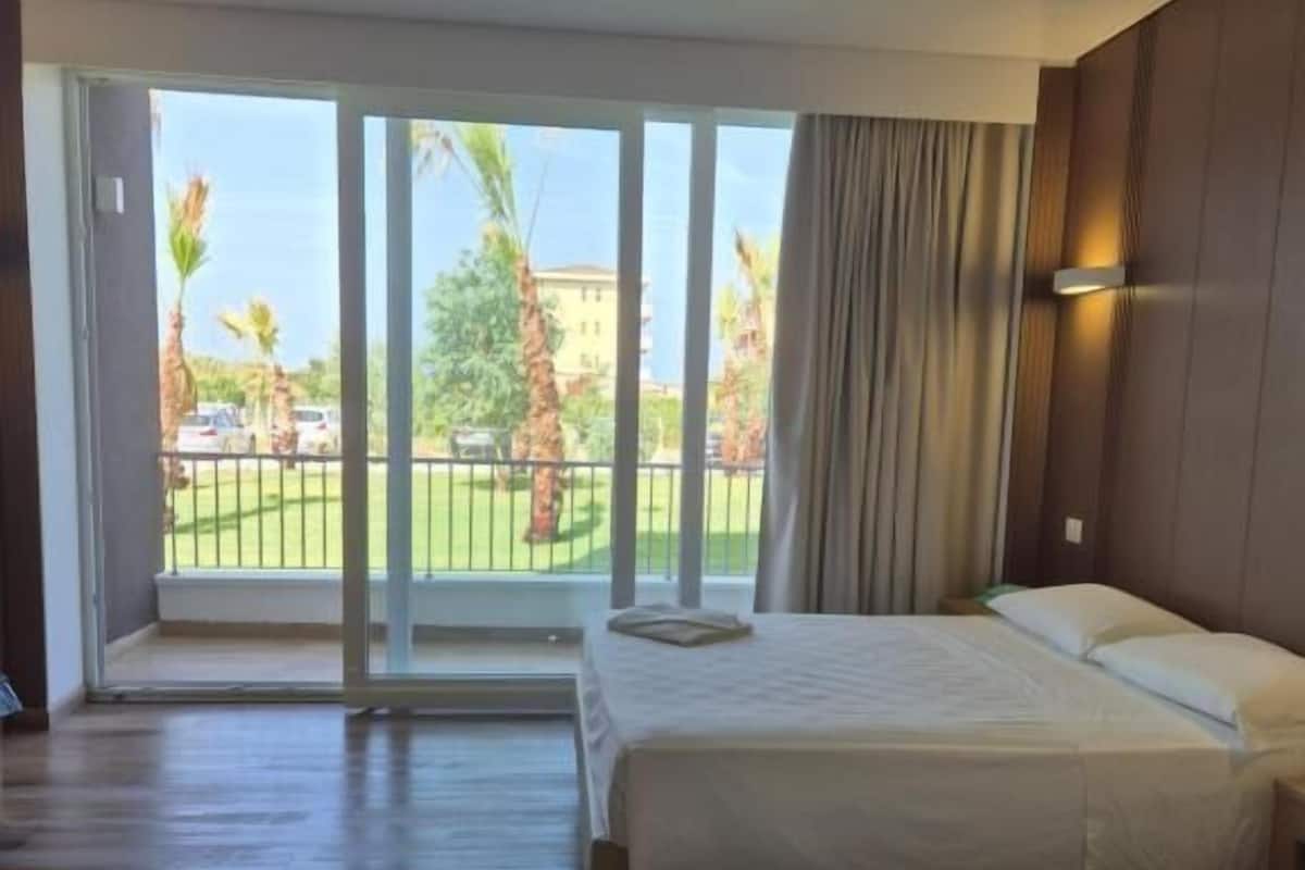 Quarto de hotel com duas camas, uma grande porta de vidro deslizante para uma varanda e vista para palmeiras e edifícios.