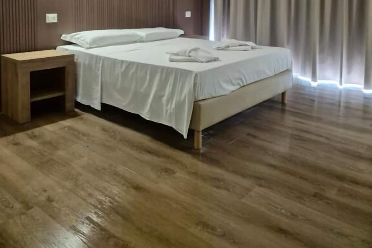 Quarto moderno com piso de madeira, cama com roupa branca, mesa de cabeceira e luminária fixada na parede.