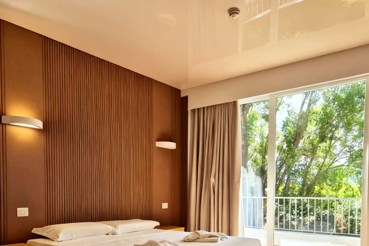 Quarto moderno com piso de madeira, uma cama grande, mesa de cabeceira e vista para uma varanda com vegetação.