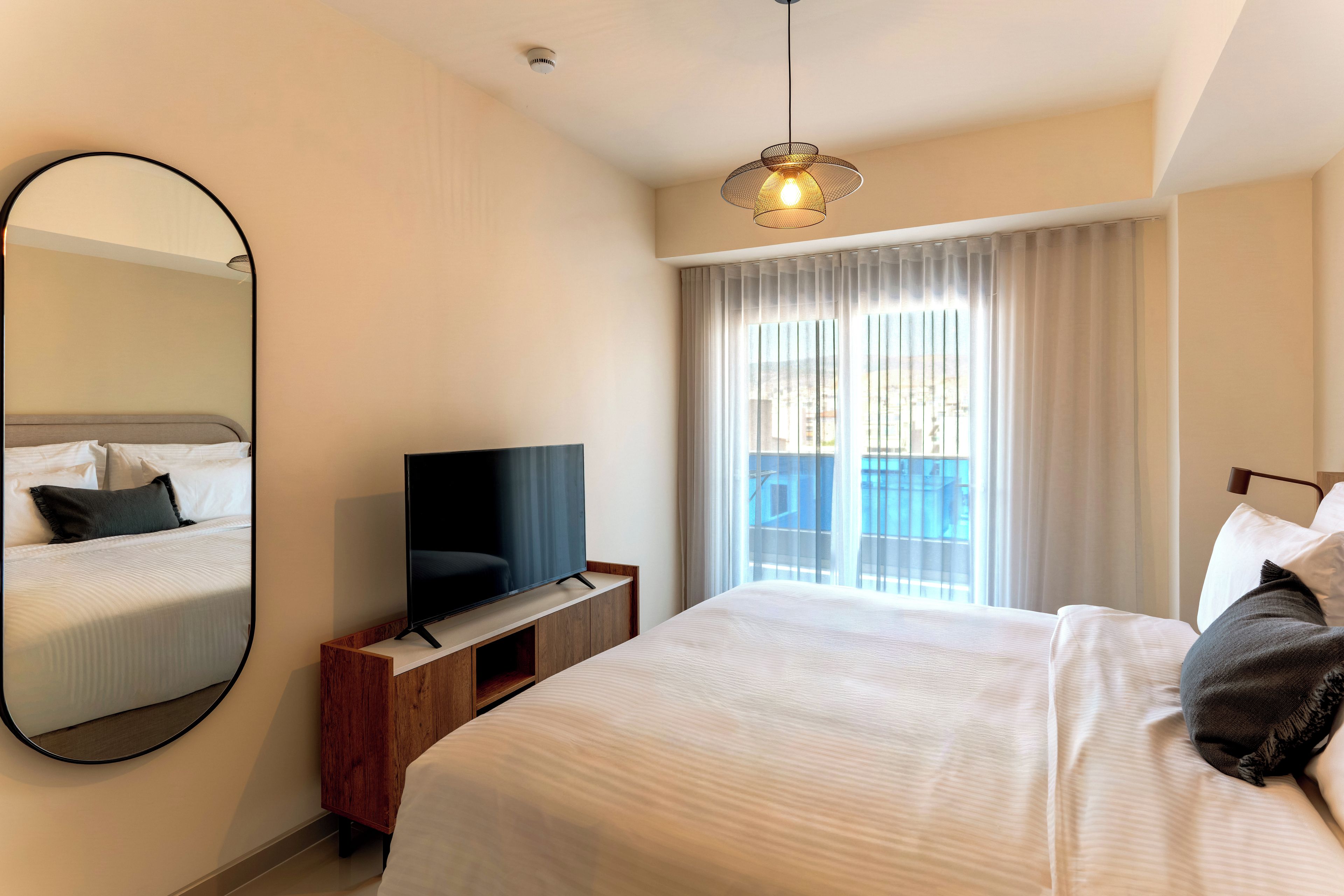 Quarto de hotel com uma cama grande, televisão de tela plana, espelho e vista para o exterior através de uma janela com cortinas.