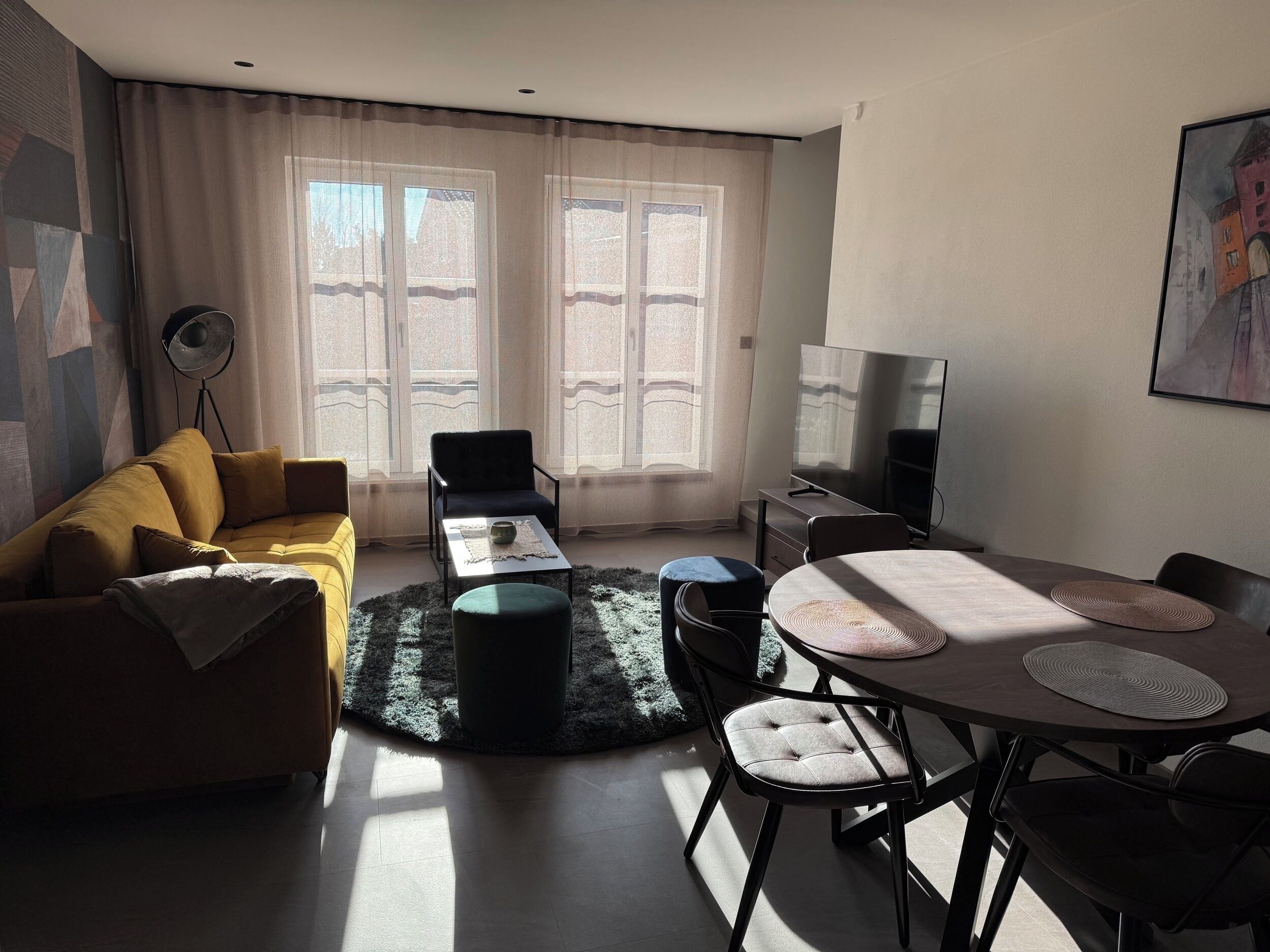 Uma sala de estar moderna com um sofá, uma mesa de centro, uma mesa de jantar com cadeiras, uma televisão e janelas grandes com cortinas.