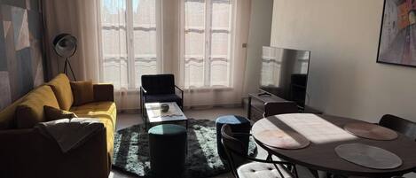 Uma sala de estar moderna com um sofá, uma mesa de centro, uma mesa de jantar com cadeiras, uma televisão e janelas grandes com cortinas.