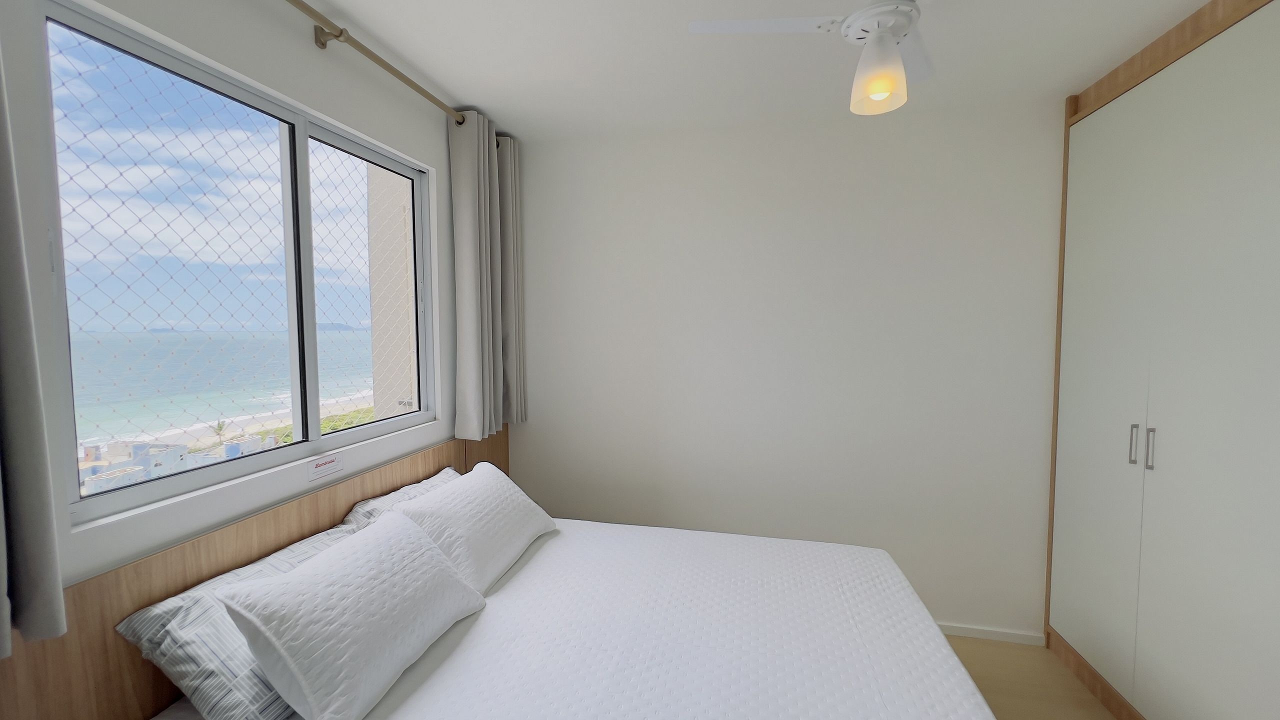 Quarto com cama, janela com cortinas, ventilador de teto e vista para o mar.