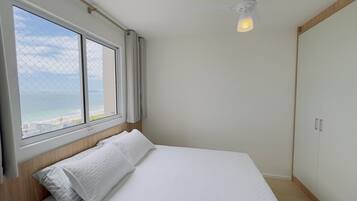 Quarto com cama, janela com cortinas, ventilador de teto e vista para o mar.