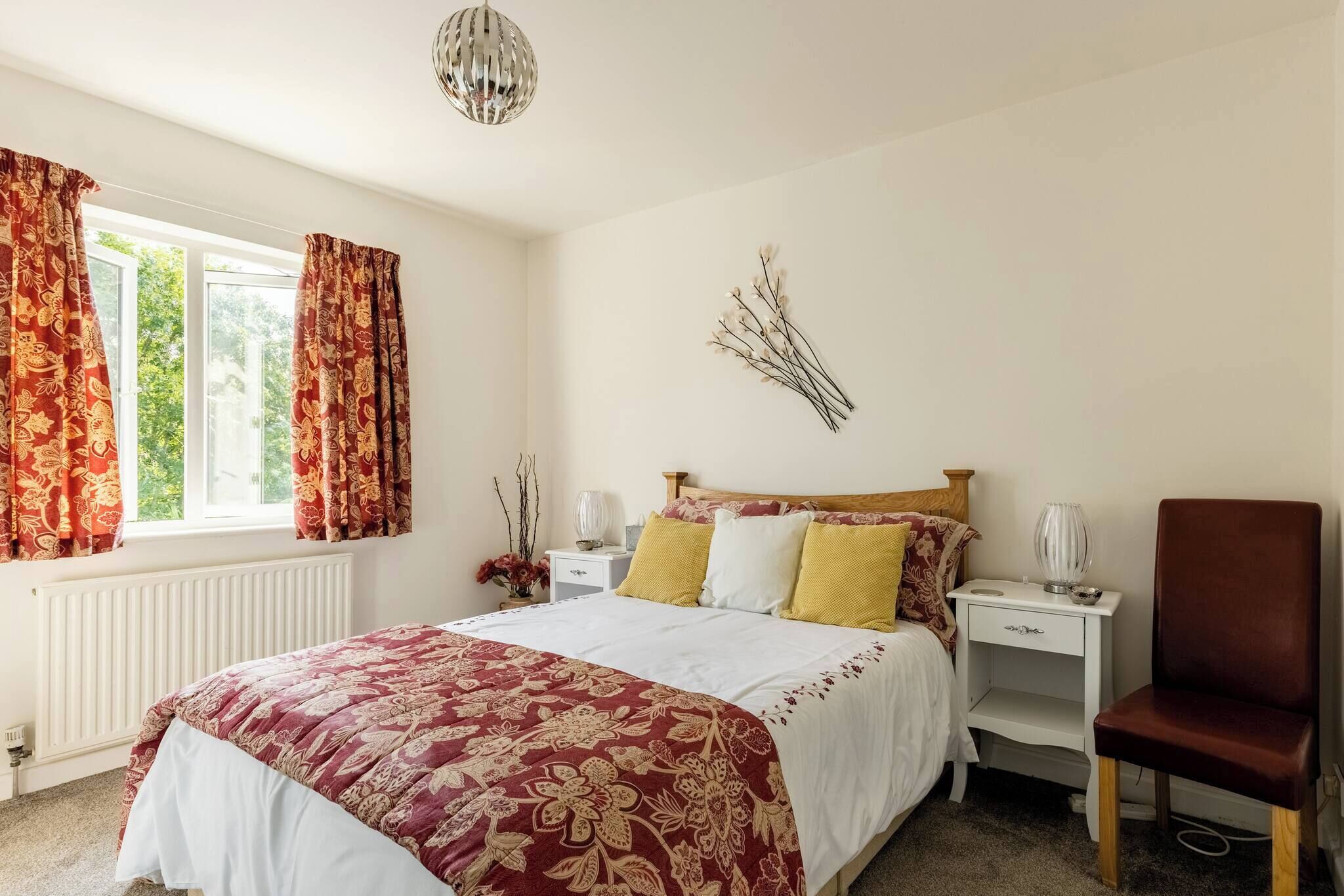 Um quarto com uma cama grande, uma mesa de cabeceira, uma cadeira, uma janela com cortinas, um radiador e um objeto decorativo na parede.