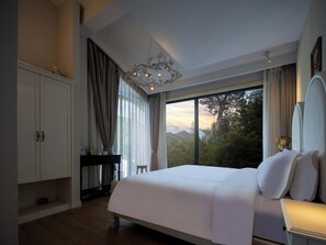 Uma cama grande com roupa de cama branca, uma mesa de cabeceira e uma escrivaninha com uma cadeira. Há uma janela ampla com cortinas, e um lustre sobre a cama.