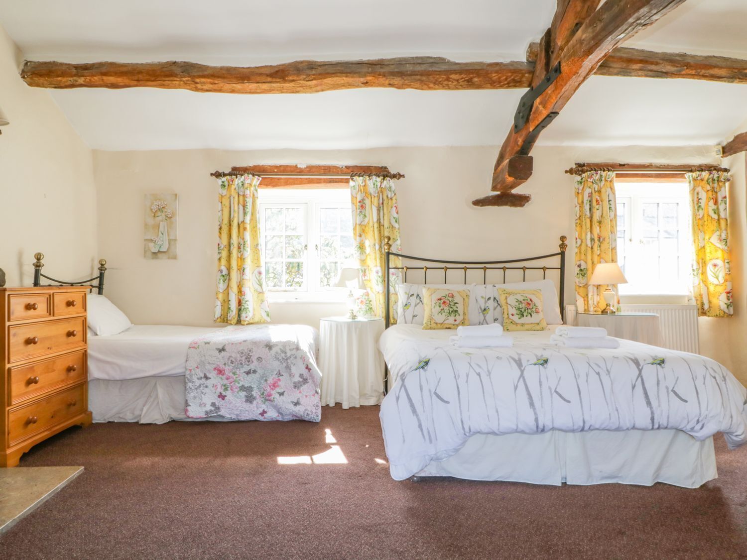 Quarto com duas camas, cómoda de madeira, cortinas florais e vigas de madeira aparente.