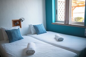 Quarto de hotel com duas camas, cada uma com roupa de cama branca e almofadas listradas em azul. Há uma janela com vista para o exterior e uma cabeceira de madeira com luminária.