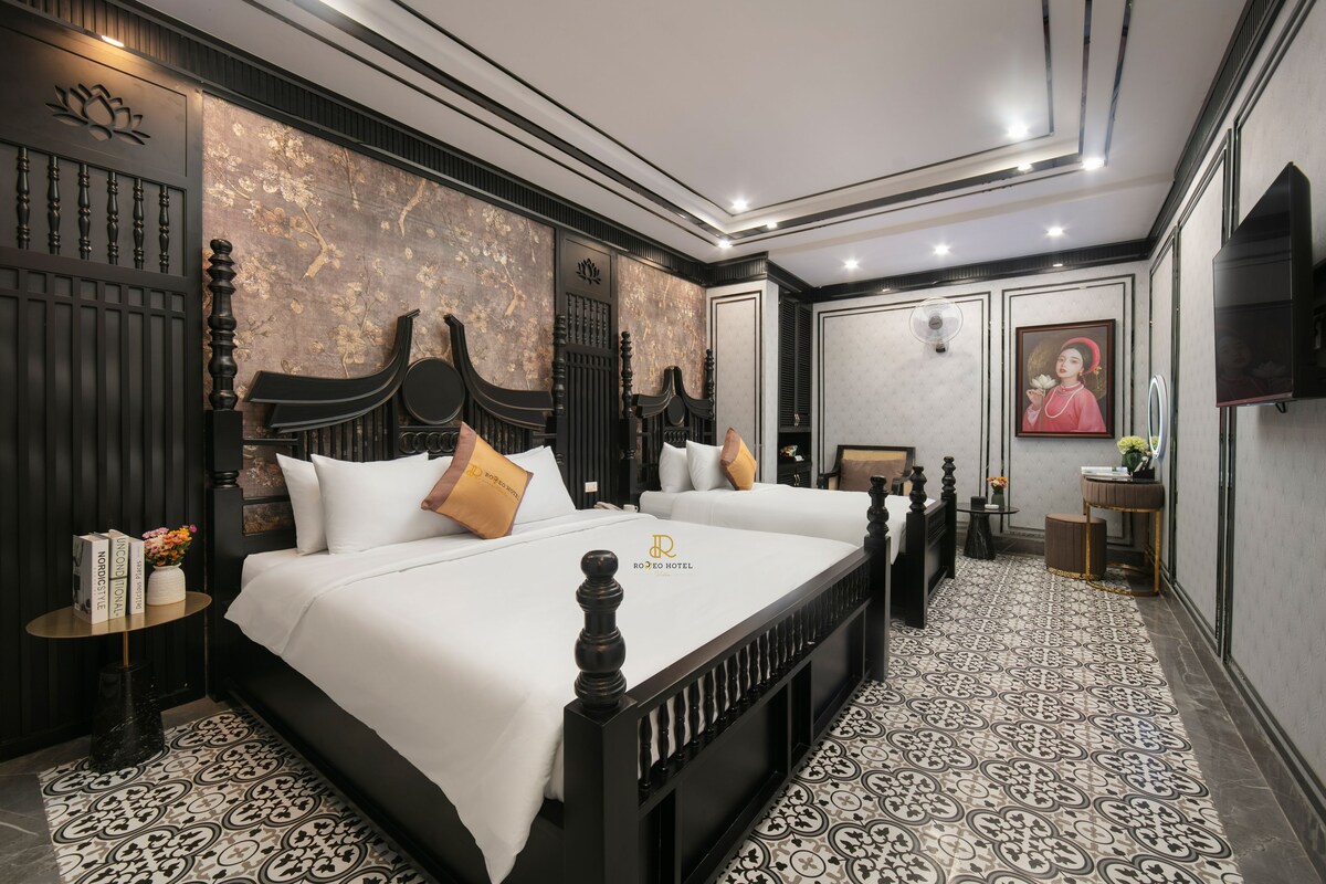 Quarto de hotel com uma cama grande, duas mesas de cabeceira, uma mesinha, uma cadeira, uma televisão e um painel decorativo.